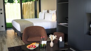 Quarto de hotel moderno com uma cama grande, um sofá, uma mesa de centro com frutas e bebidas, e vista para o exterior.