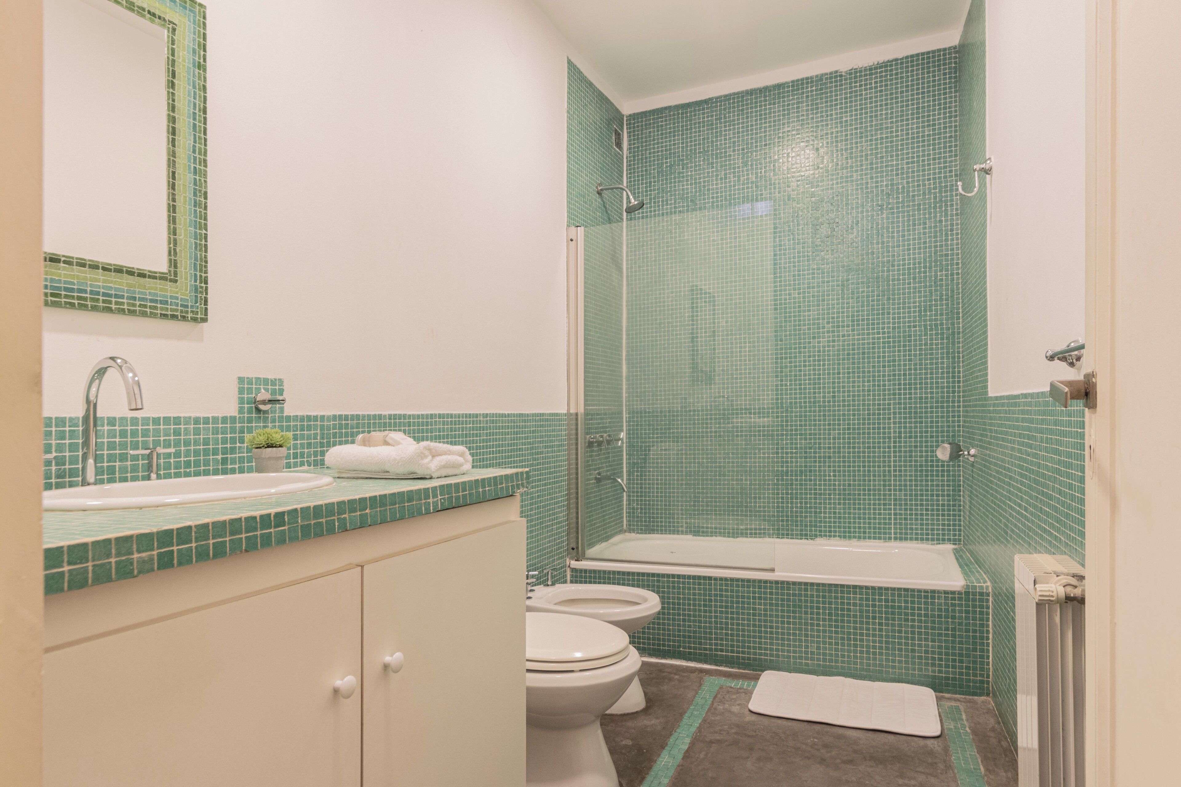 Uma casa de banho com chuveiro revestido a azulejos verdes, uma banheira branca, sanita branca e uma pia com bancada verde.