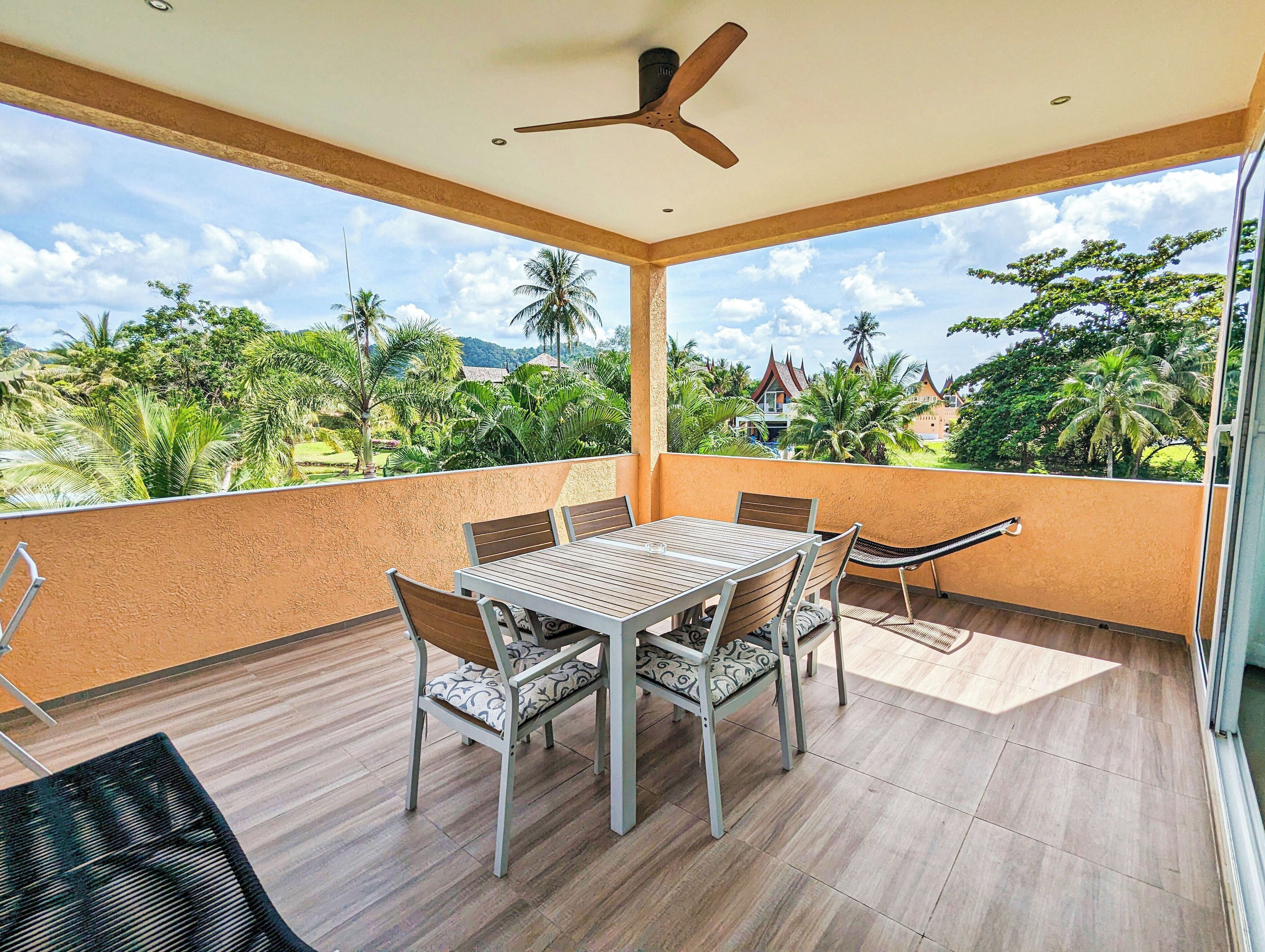 Uma varanda com mesa e cadeiras, um ventilador de teto e vista para uma paisagem tropical com palmeiras e edifícios.