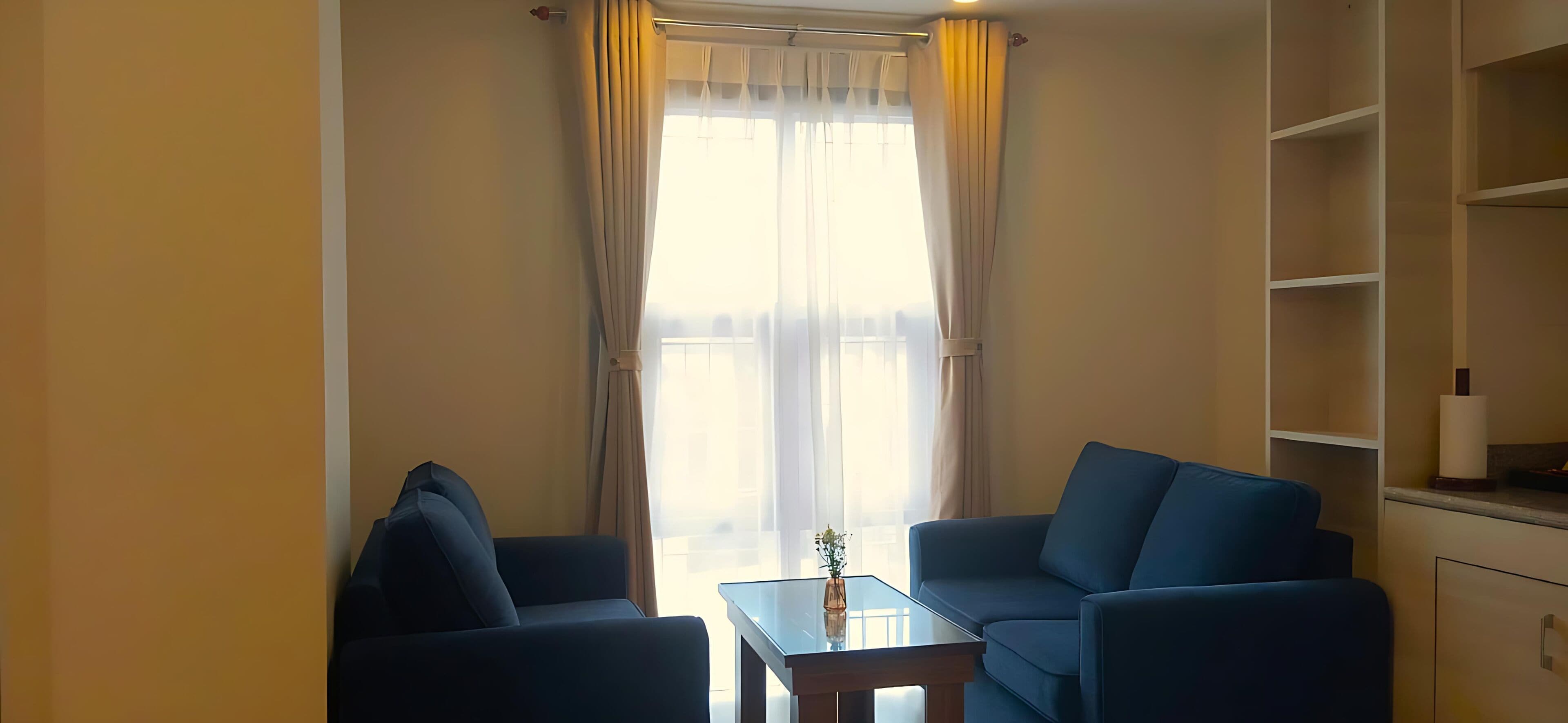 Um quarto com dois sofás azuis, uma mesa pequena com um vaso e uma janela com cortinas leves.