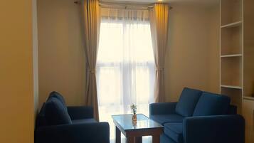 Um quarto com dois sofás azuis, uma mesa pequena com um vaso e uma janela com cortinas leves.