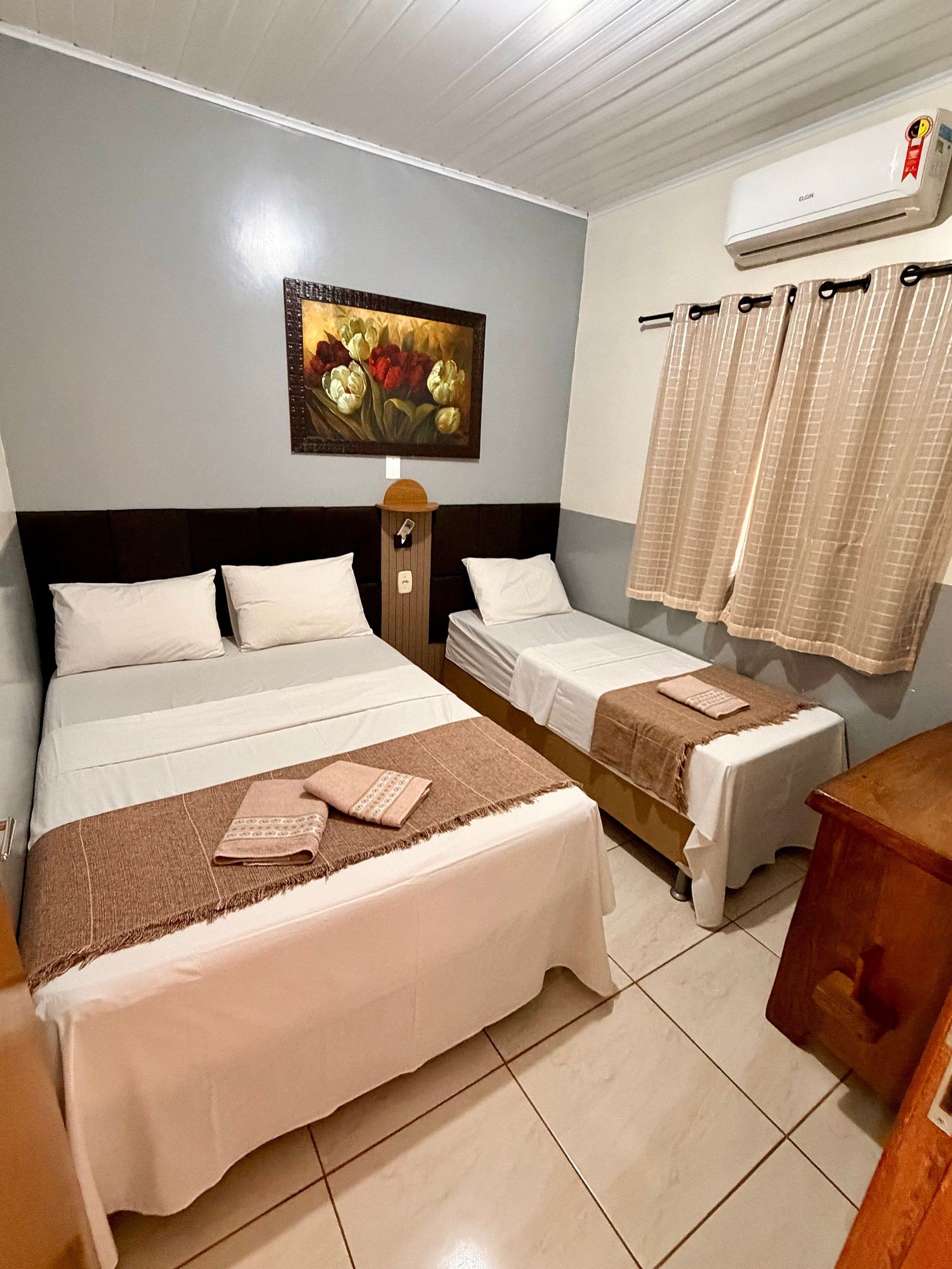 Quarto de hotel com duas camas, um quadro na parede, aparelho de ar condicionado e janela com cortinas.