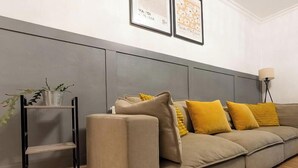 Um sofá em tons neutros com almofadas amarelas, uma pequena mesa lateral com uma planta e um abajur.
