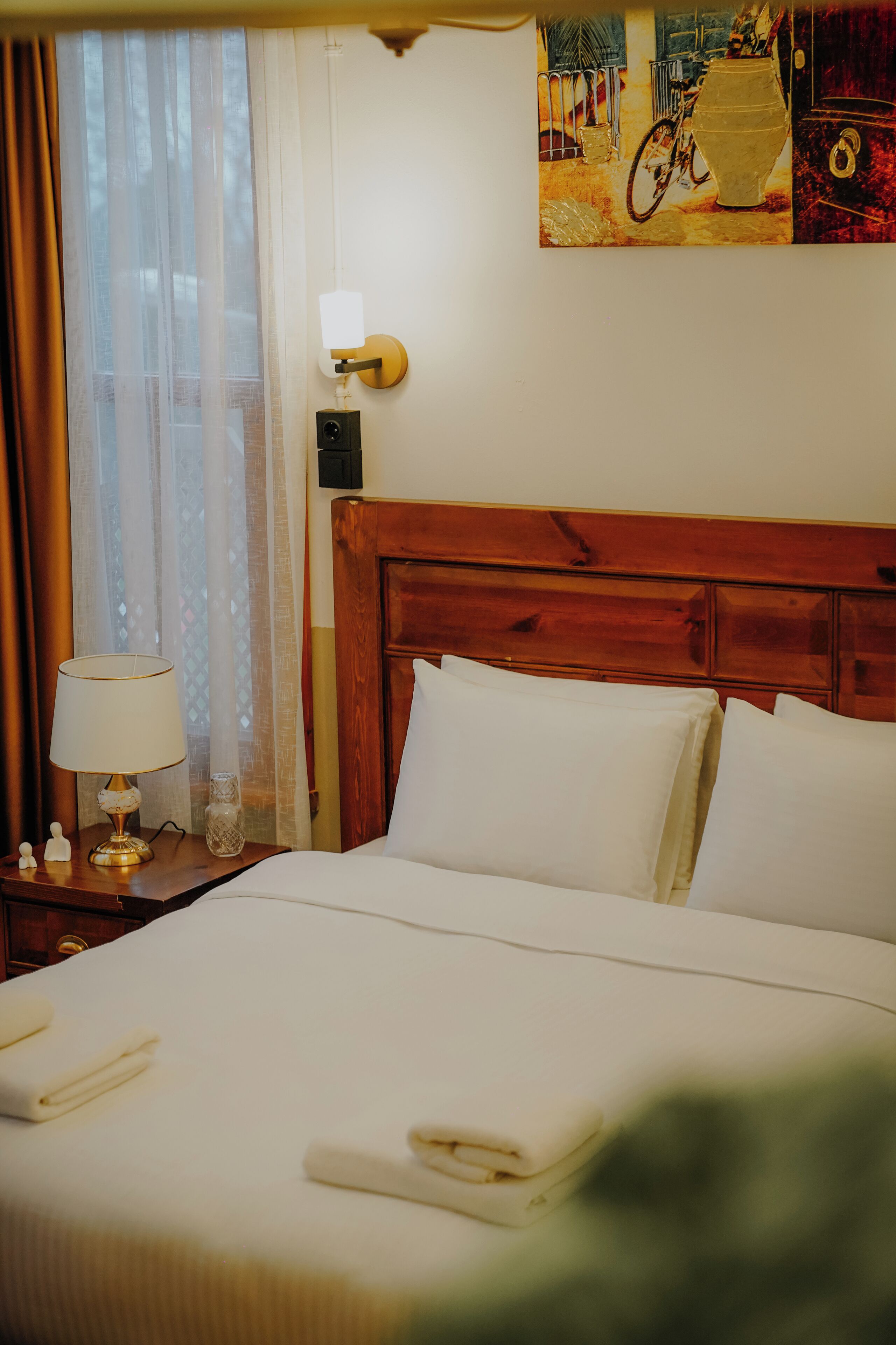 Quarto de hotel com cabeceira de madeira, cama com lençóis brancos, mesa de cabeceira com abajur e um quadro na parede.