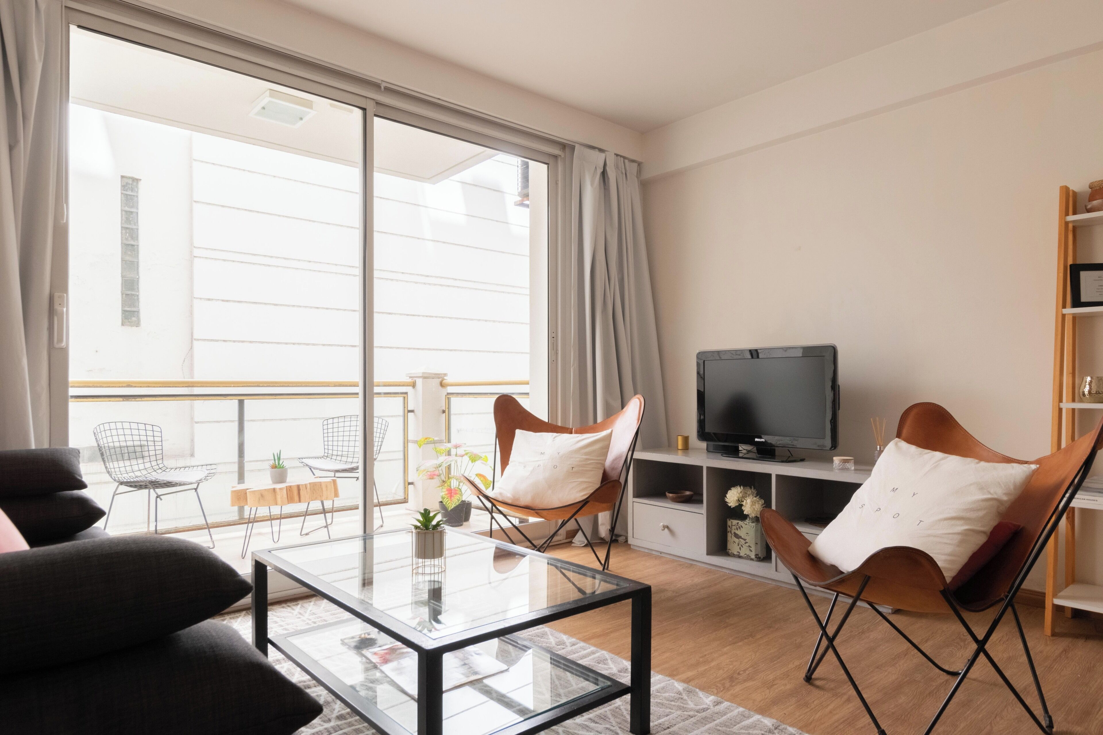 Uma sala de estar moderna com uma mesa de centro de vidro, poltronas de couro, uma televisão de tela plana e uma varanda com mobiliário para áreas externas.
