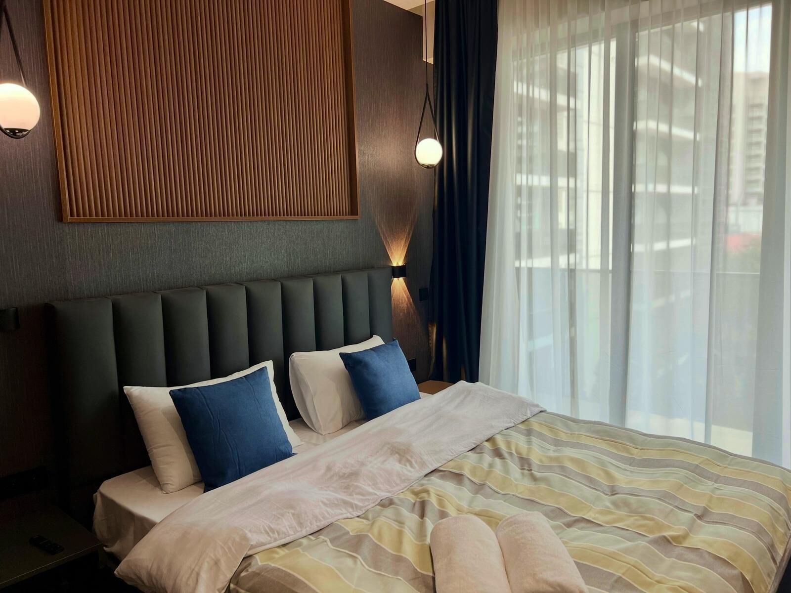 Quarto de hotel com uma cama grande, cabeceira de madeira, janela com cortinas e luminária fixada na parede.