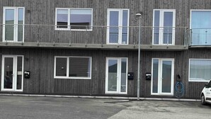 Um edifício de vários andares com fachada cinza, diversas janelas e uma área de estacionamento com um carro branco.