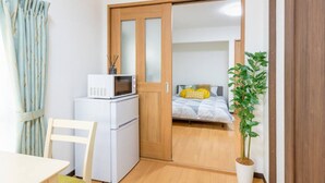 Quarto compacto com cama, micro-ondas, mini-geladeira, porta de madeira e uma planta em vaso.
