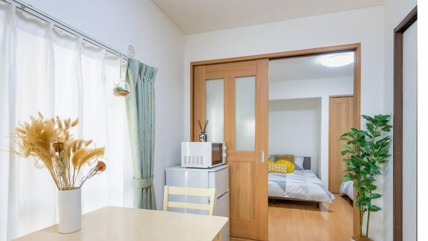 Quarto compacto com cama, micro-ondas, uma mesinha com um vaso de flores secas e uma planta em vaso.