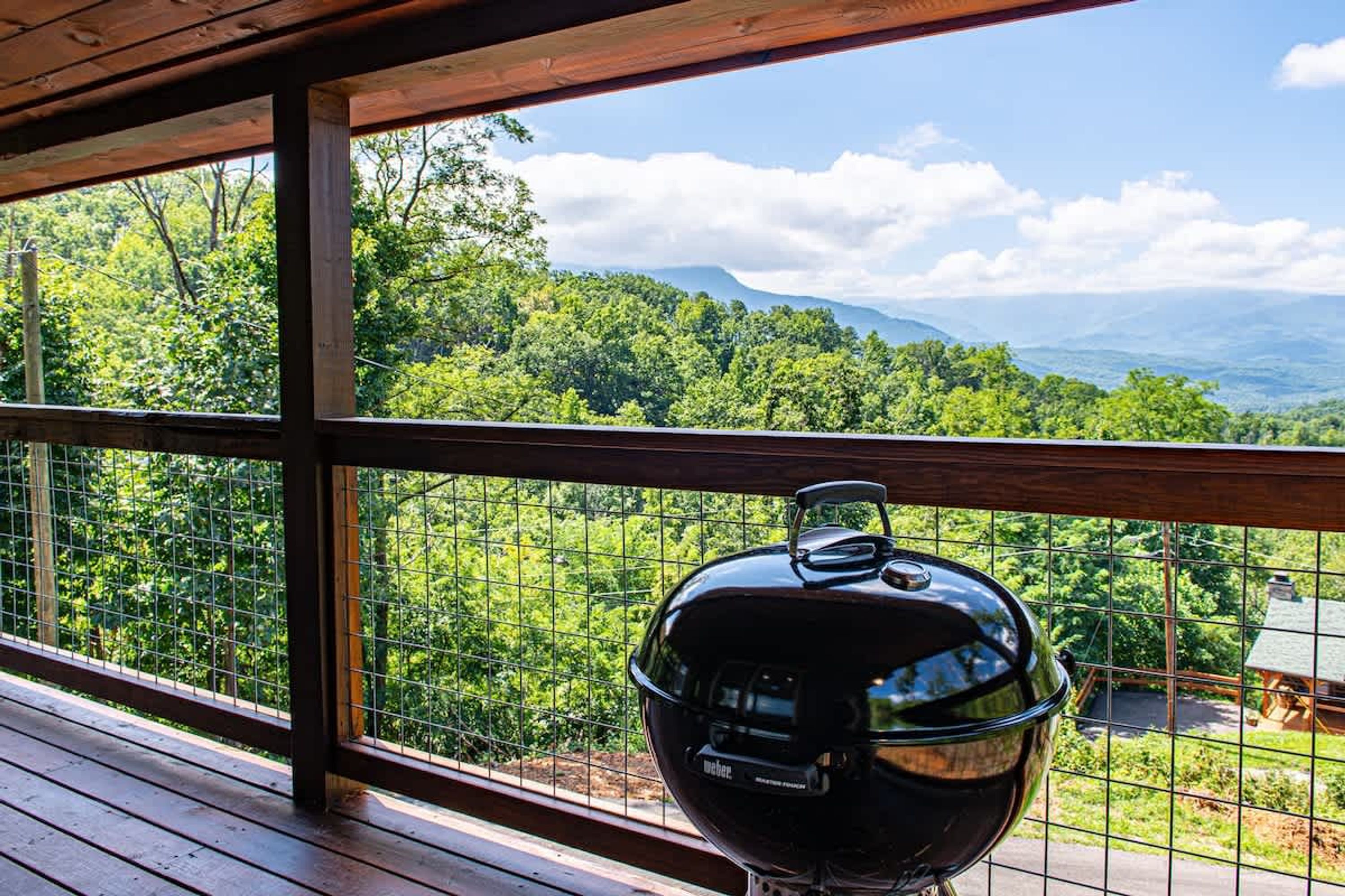 Vista de um deck de madeira com churrasqueira e cercado de arame, com vista para uma paisagem florestal.