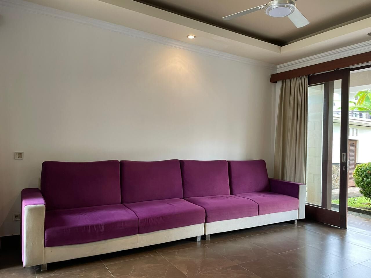 Um sofá roxo num quarto com ventoinha no teto, porta de correr de vidro e vista para o verde lá fora.