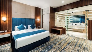 Quarto de hotel moderno com uma cama grande, mesa de cabeceira, casa de banho com divisórias de vidro e uma parede de destaque em madeira.