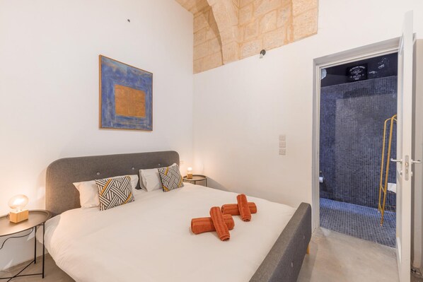 Quarto de hotel com uma cama grande, uma mesa de cabeceira com abajur, área de chuveiro com parede azulejada e uma obra de arte emoldurada na parede.