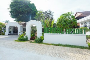 Placa do Herbal Resort, com entrada pavimentada e vegetação ao redor.