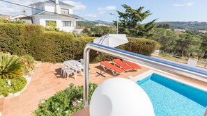 Piscina no terraço com cadeiras de sol vermelhas, uma mesa e cadeiras, cercada por vegetação e vista para uma casa e colinas.