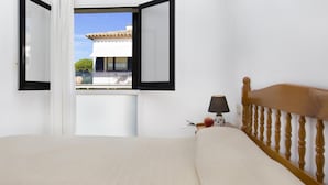 Quarto com cama, mesa de cabeceira com abajur e janela com cortina aberta.