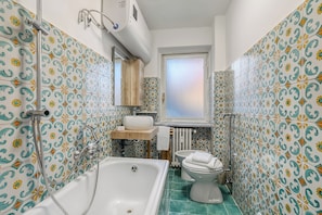 Casa de banho com banheira, sanita e lavatório, apresentando azulejos estampados e uma janela.