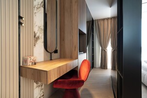 Quarto de hotel moderno com uma escrivaninha de madeira, uma cadeira vermelha, um espelho grande e vista para o exterior através de cortinas.