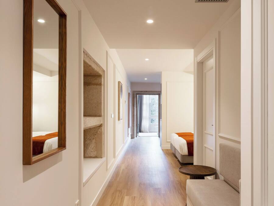 Um corredor com piso de madeira, uma cama com colchão branco e roupa de cama laranja, e um espelho grande na parede à esquerda.