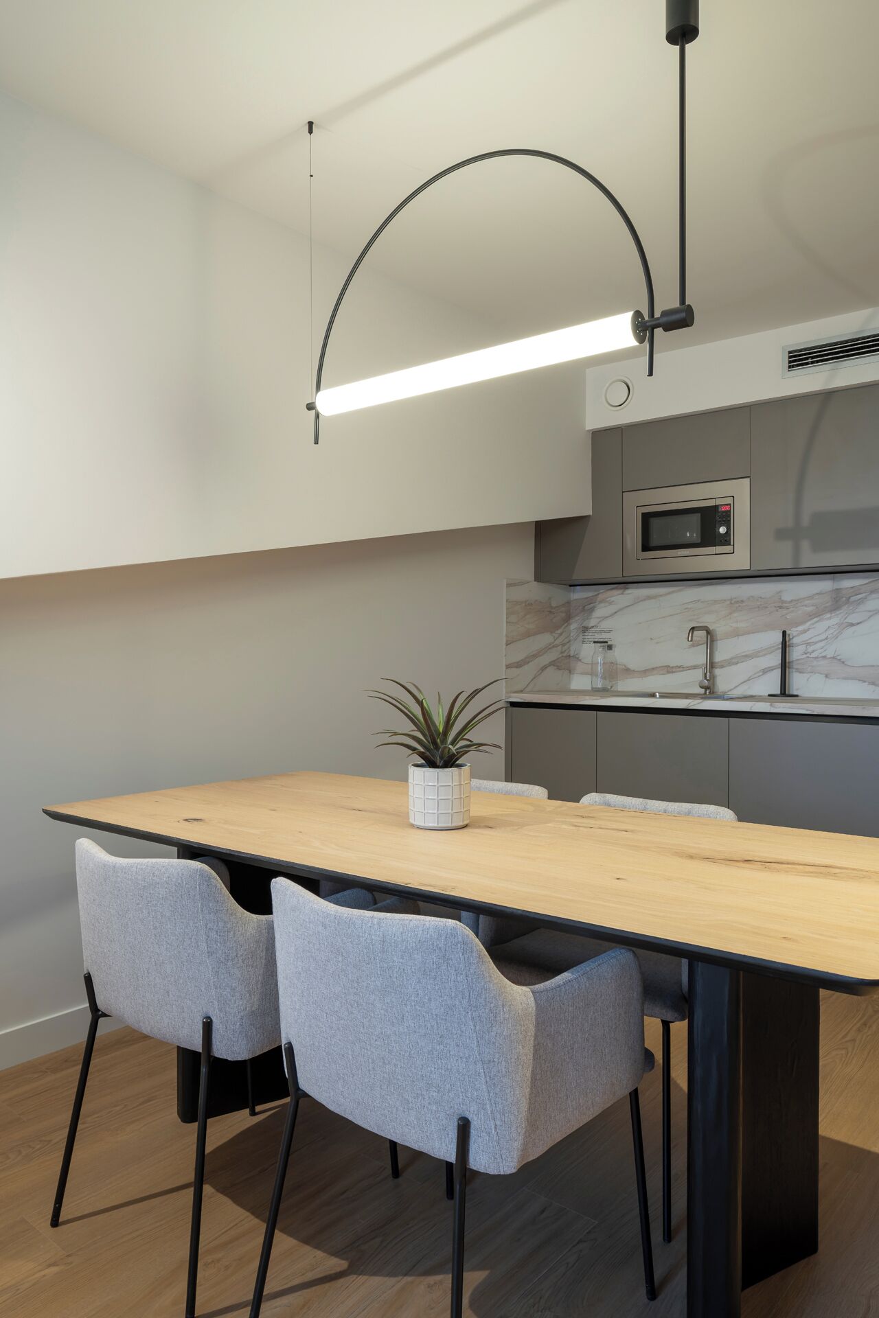Uma cozinha moderna com uma mesa de madeira, duas cadeiras cinzentas, um vaso com planta e um micro-ondas embutido.