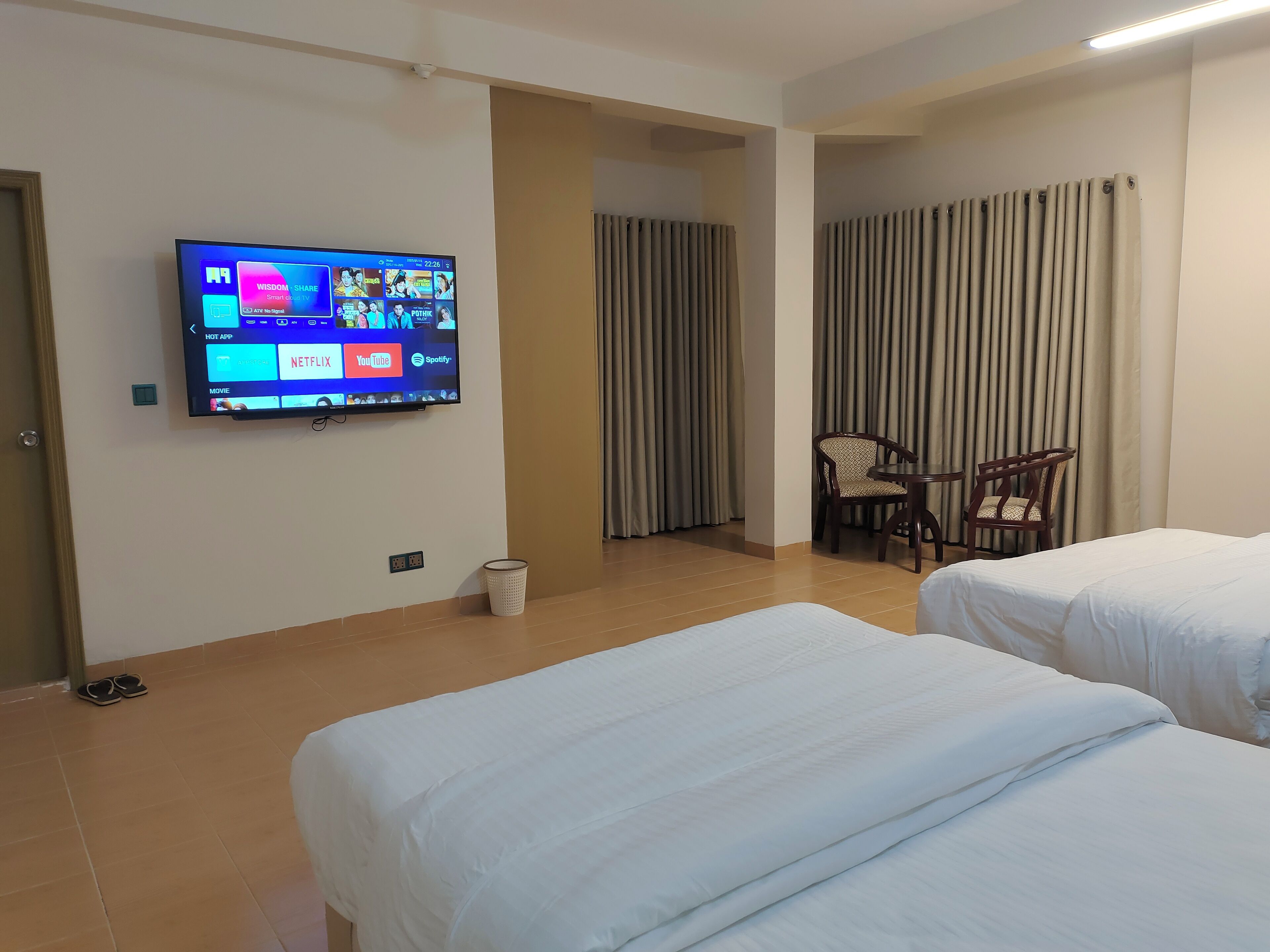 Quarto de hotel com televisão exibindo diversos aplicativos de streaming, duas camas, uma mesinha com cadeiras e uma cortina.