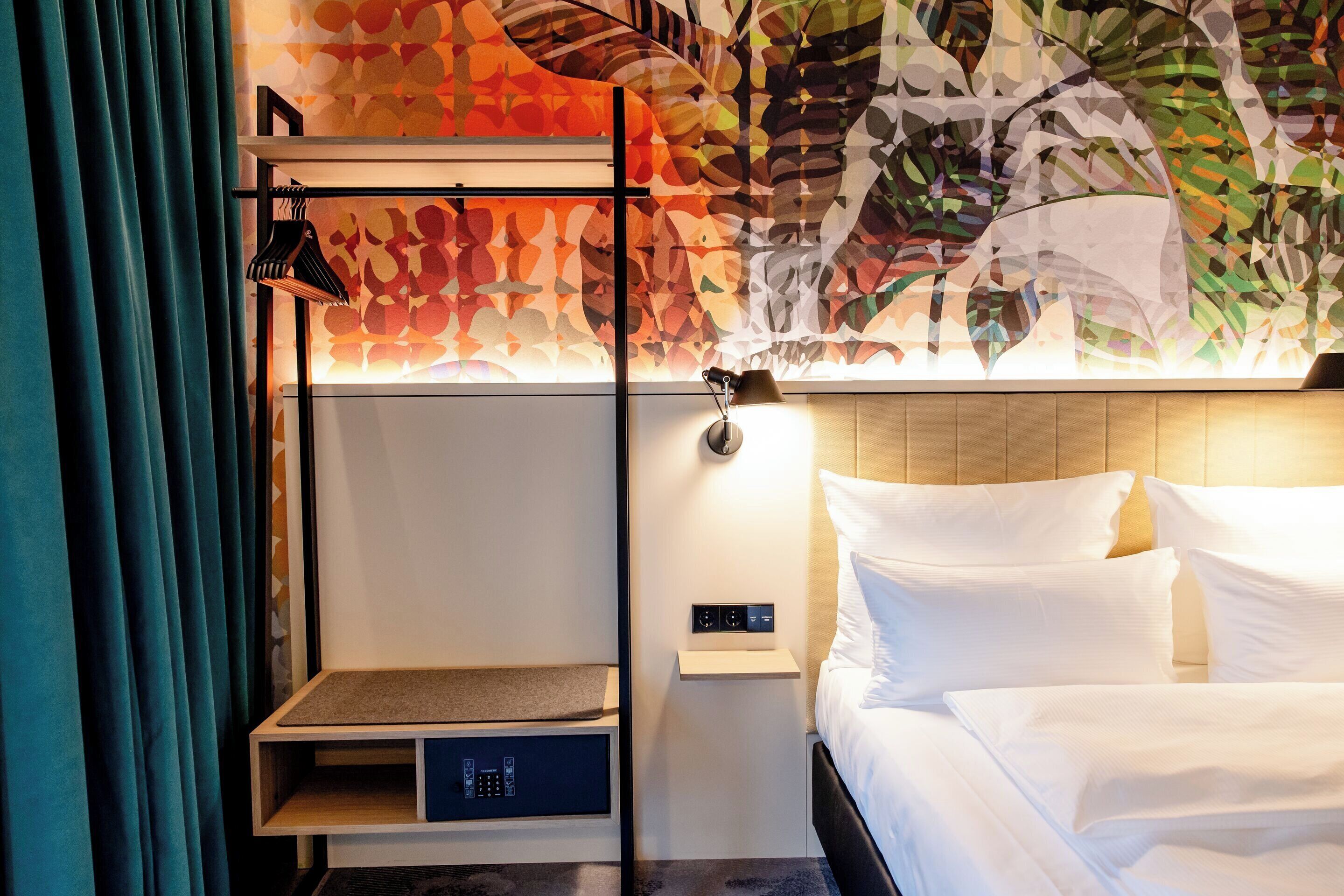 Quarto de hotel com cama, mesa de cabeceira, luminária, prateleira e parede com padrão de folhas.