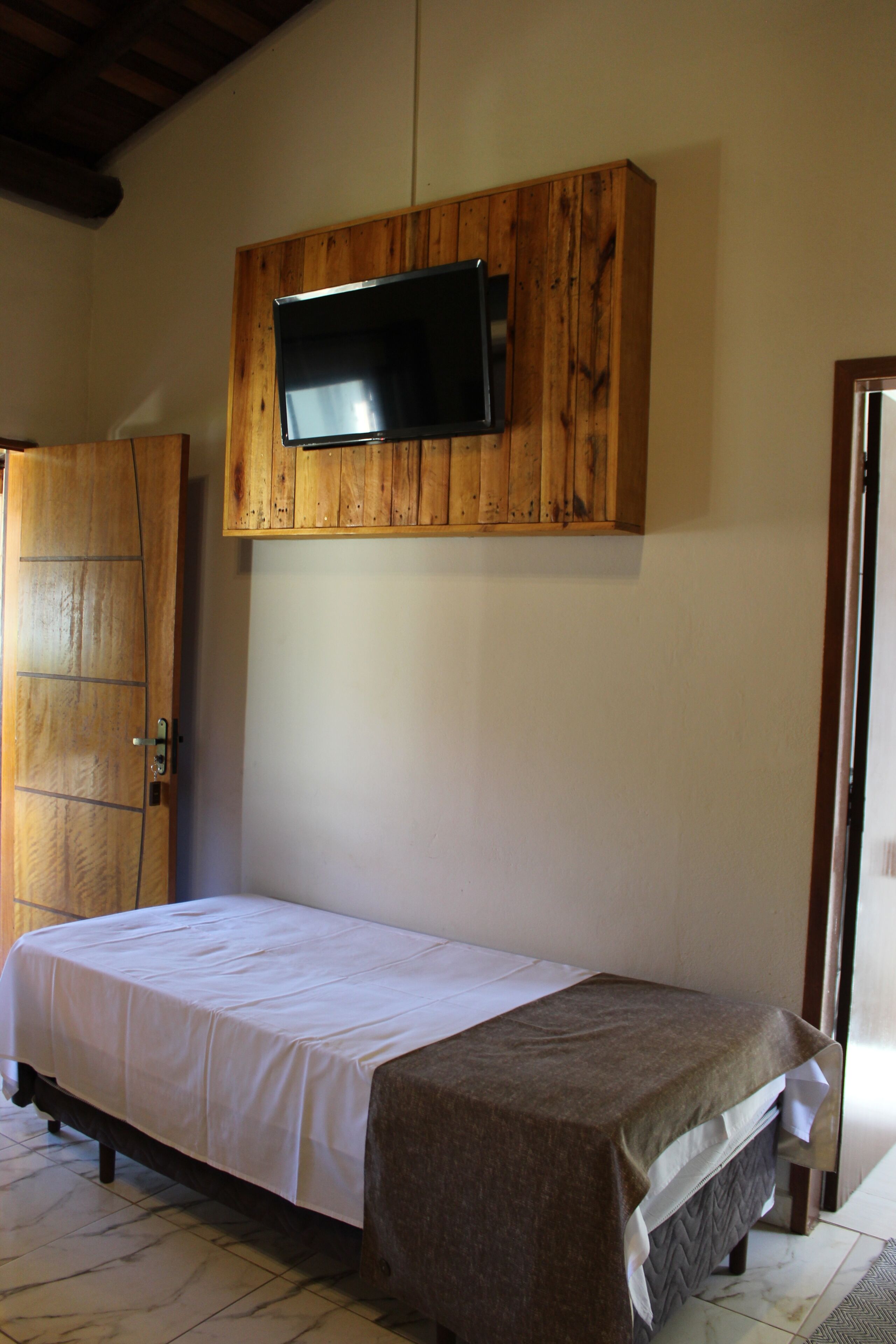 Uma cama de solteiro com lençol branco e manta marrom, um armário de madeira com televisão de tela plana, e uma porta à esquerda.