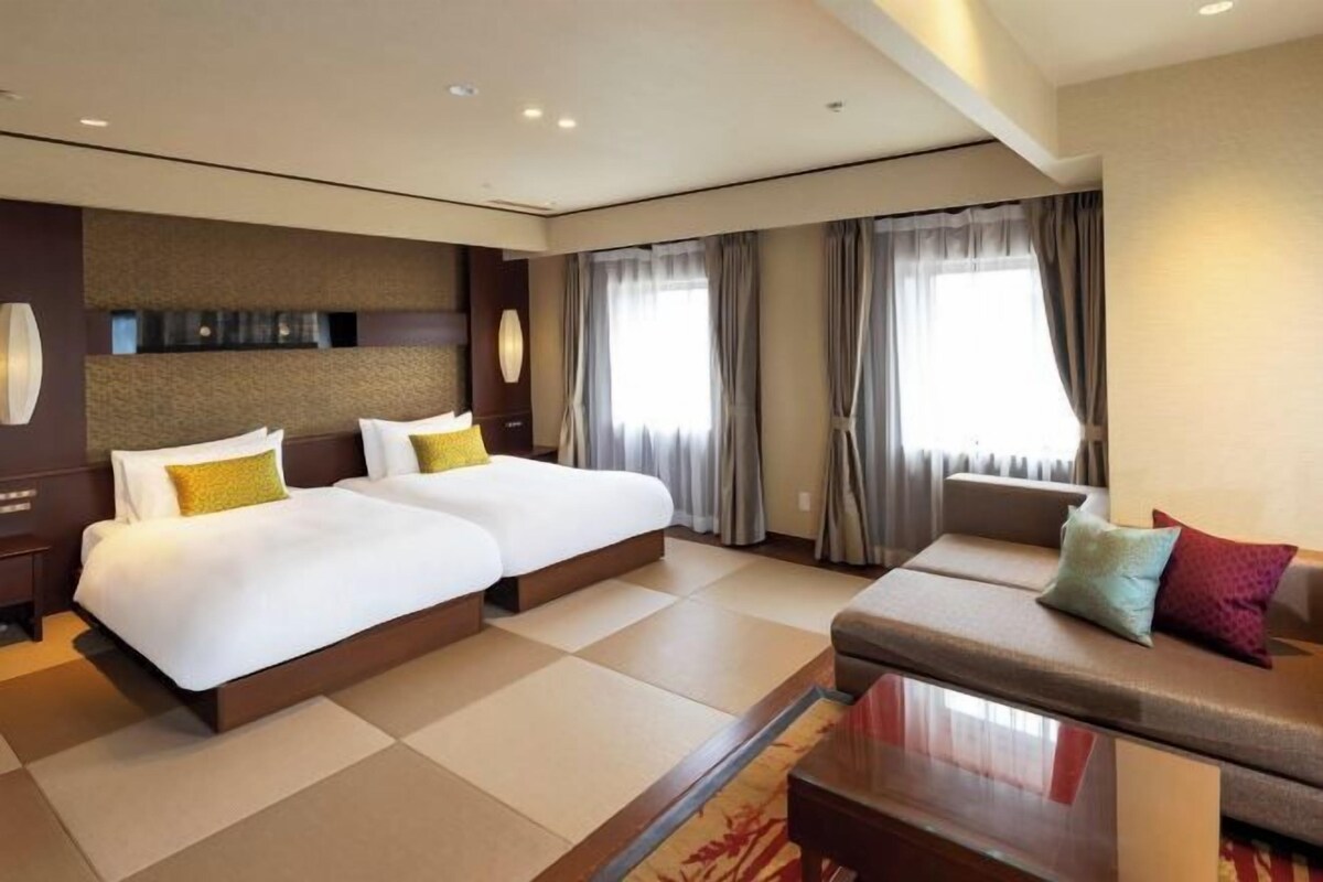 Quarto de hotel com duas camas, um sofá, uma mesa de centro e uma janela grande com cortinas.