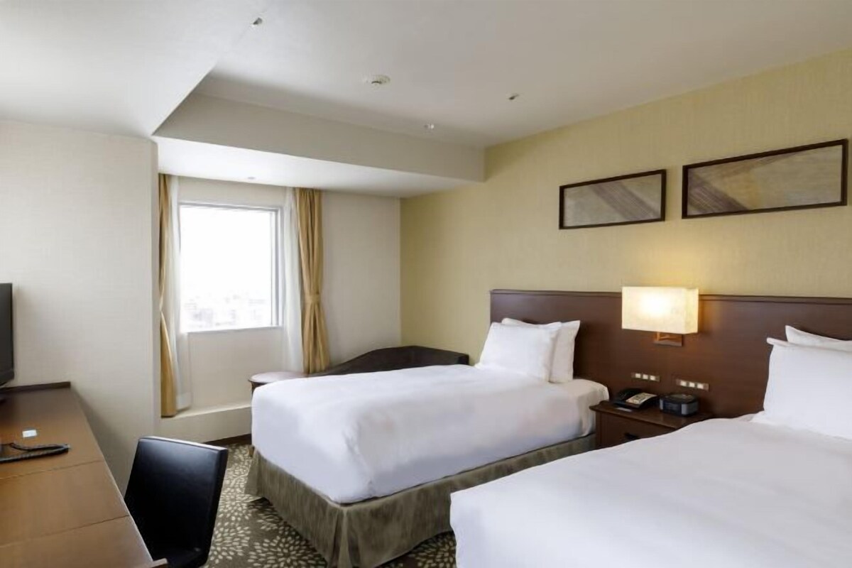 Quarto de hotel com duas camas, uma escrivaninha, uma cadeira, televisão e janela com cortinas.