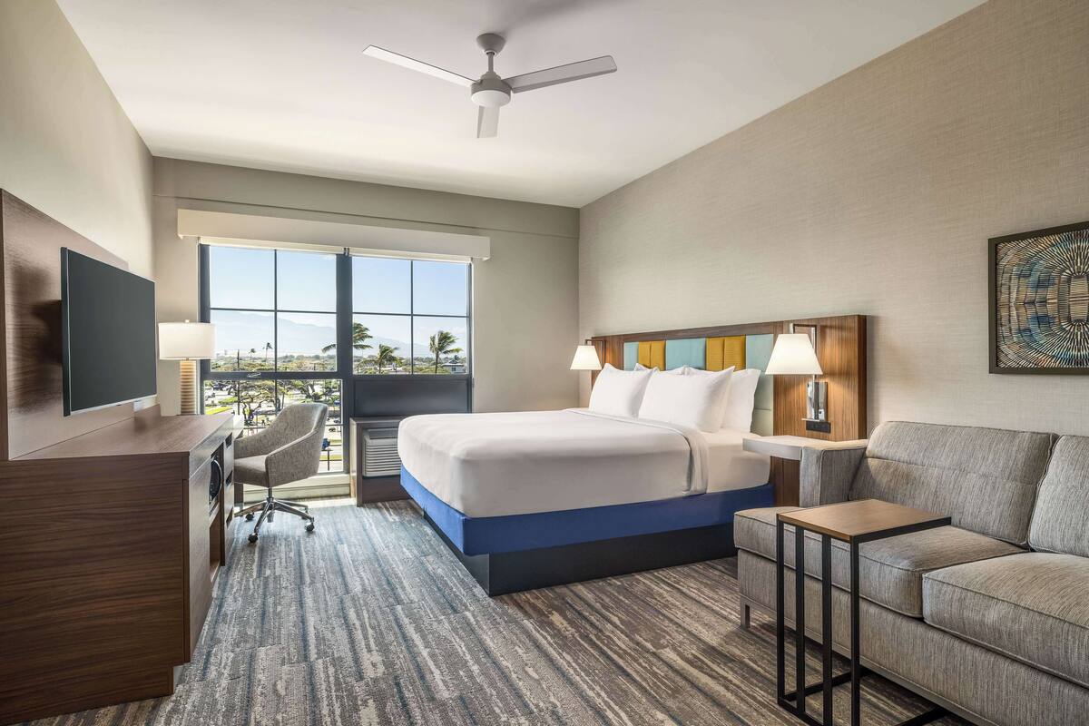Quarto de hotel com cama, escrivaninha, poltrona, sofá, ventilador de teto e uma janela grande com vista.