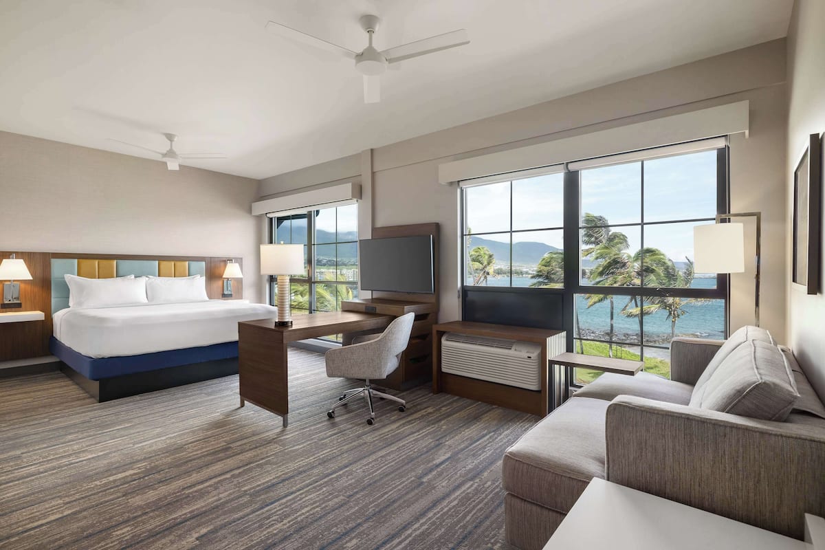 Quarto de hotel com cama, escrivaninha, cadeira e uma janela grande com vista para o mar.