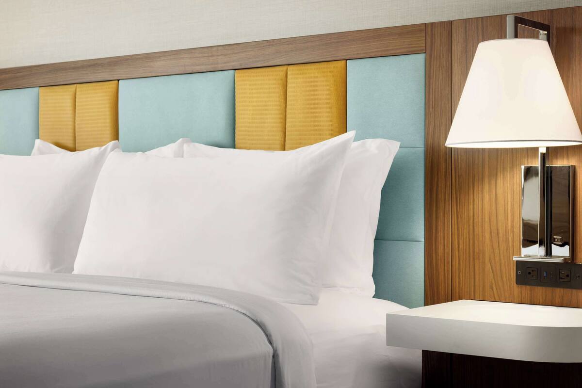 Quarto de hotel com cama, almofadas brancas, cabeceira de madeira, mesa de cabeceira e abajur.