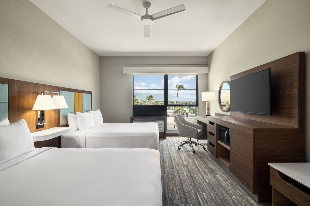 Quarto de hotel com duas camas, uma escrivaninha, televisão e uma janela grande com vista para palmeiras.