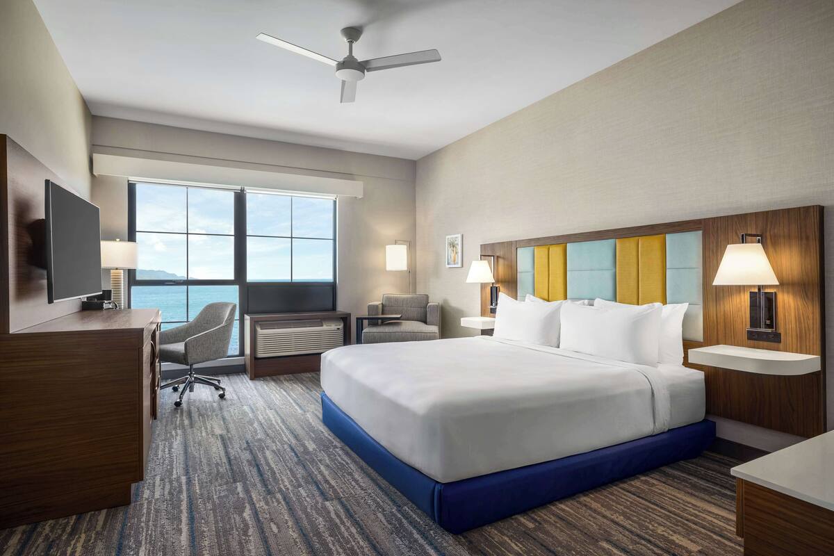 Quarto de hotel com uma cama grande, uma escrivaninha, uma cadeira, uma televisão e vista para o mar.