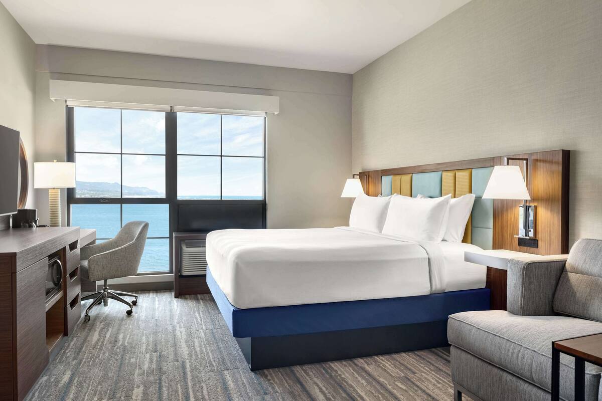 Quarto de hotel com cama grande, escrivaninha, cadeira e vista para o mar.