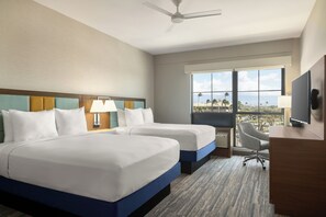 Quarto de hotel com duas camas, uma escrivaninha, uma cadeira, televisão e uma grande janela com vista para palmeiras e o oceano.
