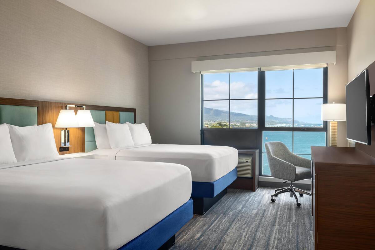 Quarto de hotel com duas camas, uma mesa, uma cadeira e uma grande janela com vista para o mar e montanhas.