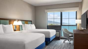 Quarto de hotel com duas camas, uma mesa, uma cadeira e uma grande janela com vista para o mar e montanhas.