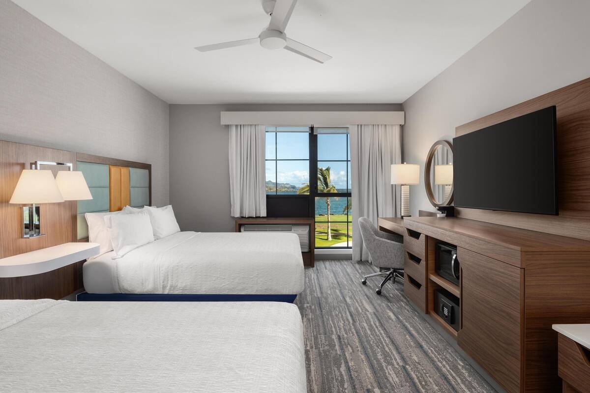 Quarto de hotel com duas camas, uma escrivaninha, televisão, ventoinha no teto e janela com vista para o exterior.