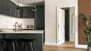 Uma cozinha moderna com armários escuros, bancada de mármore e piso de madeira. Há dois taburetes pretos e um vaso com planta.