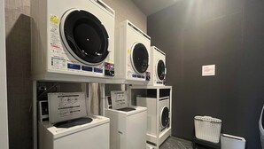 Uma lavanderia com várias máquinas de lavar e secar, chão de azulejos e um cesto para roupa.