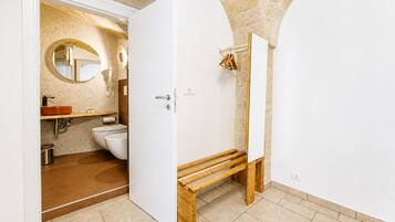 Casa de banho com um banco de madeira, uma sanita, uma pia com espelho e um porta-toalhas.