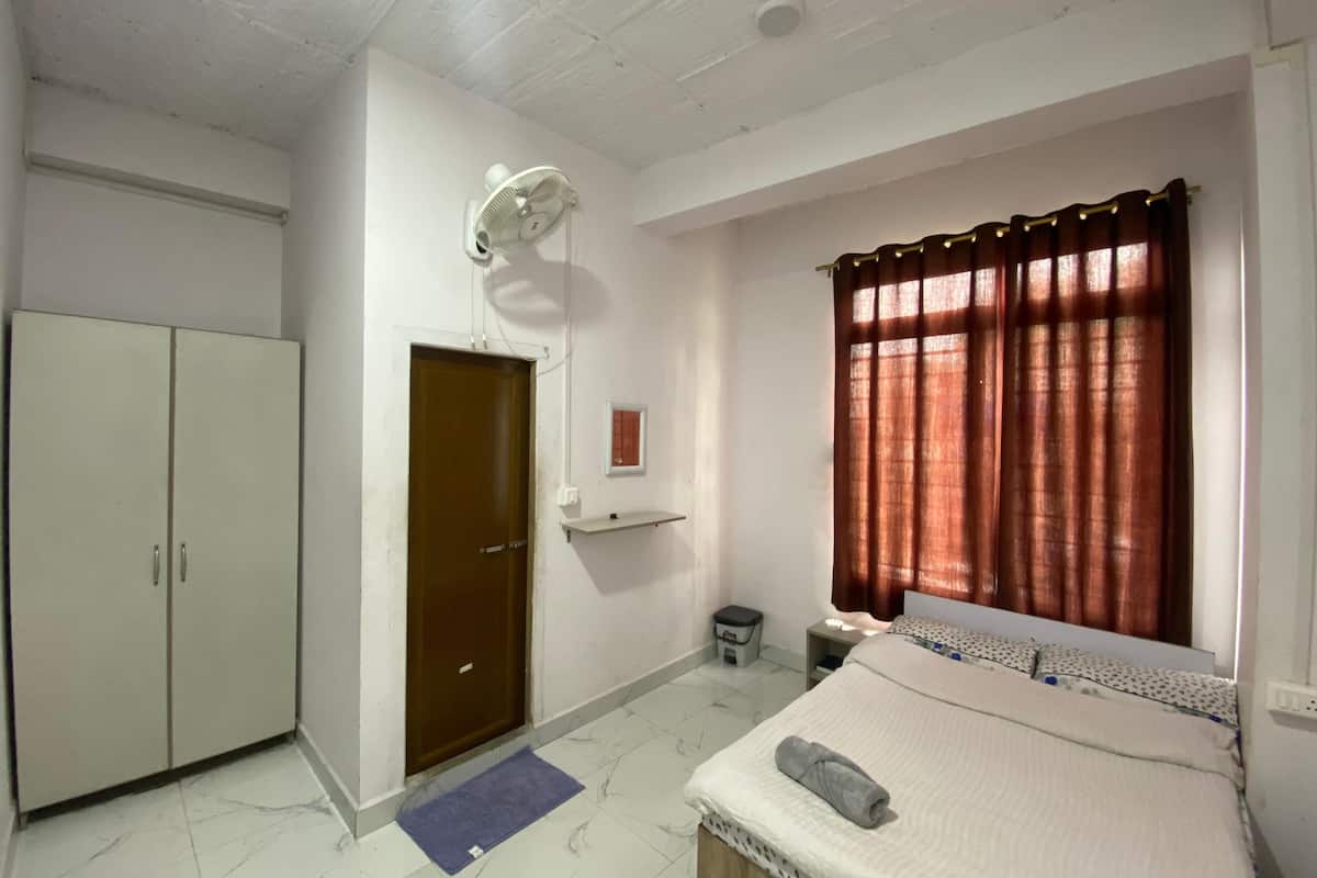 Quarto com cama, guarda-roupa, porta, pequena janela e ventoinha fixada na parede.