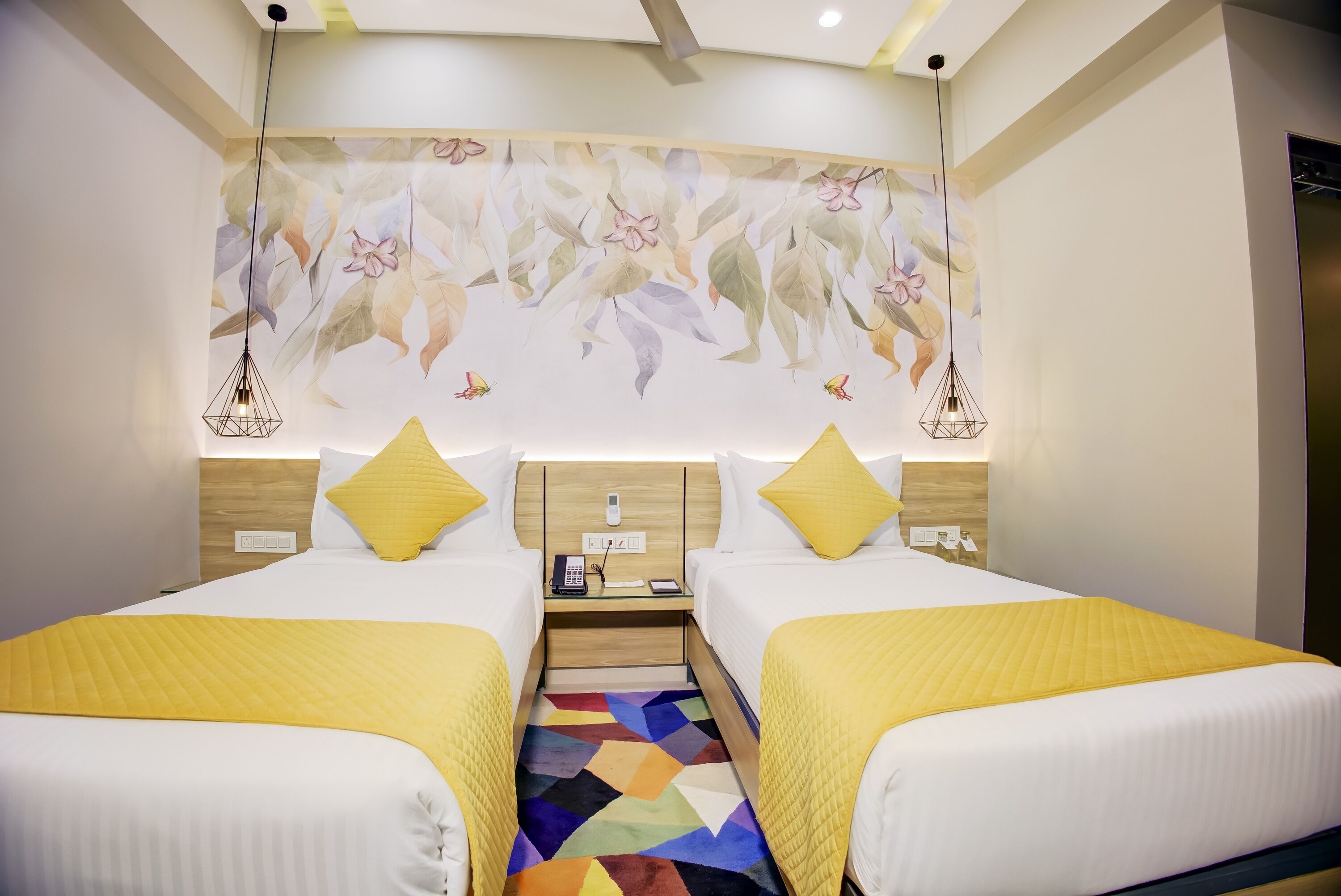 Quarto de hotel com duas camas, um tapete geométrico colorido, um mural floral na parede e um ventilador de teto.