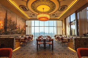 Uma área de jantar luxuosa, com um grande candelabro, teto ricamente decorado e vista para a cidade através de amplas janelas.