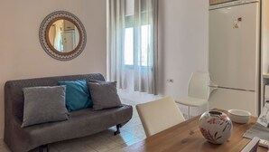 Uma sala moderna com um sofá cinzento, uma mesa de jantar de madeira, uma cadeira branca e uma cozinha com um frigorífico branco e um vaso na mesa.