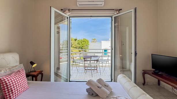 Quarto de hotel com cama, televisão, varanda com mesa e cadeiras e vista para o exterior.