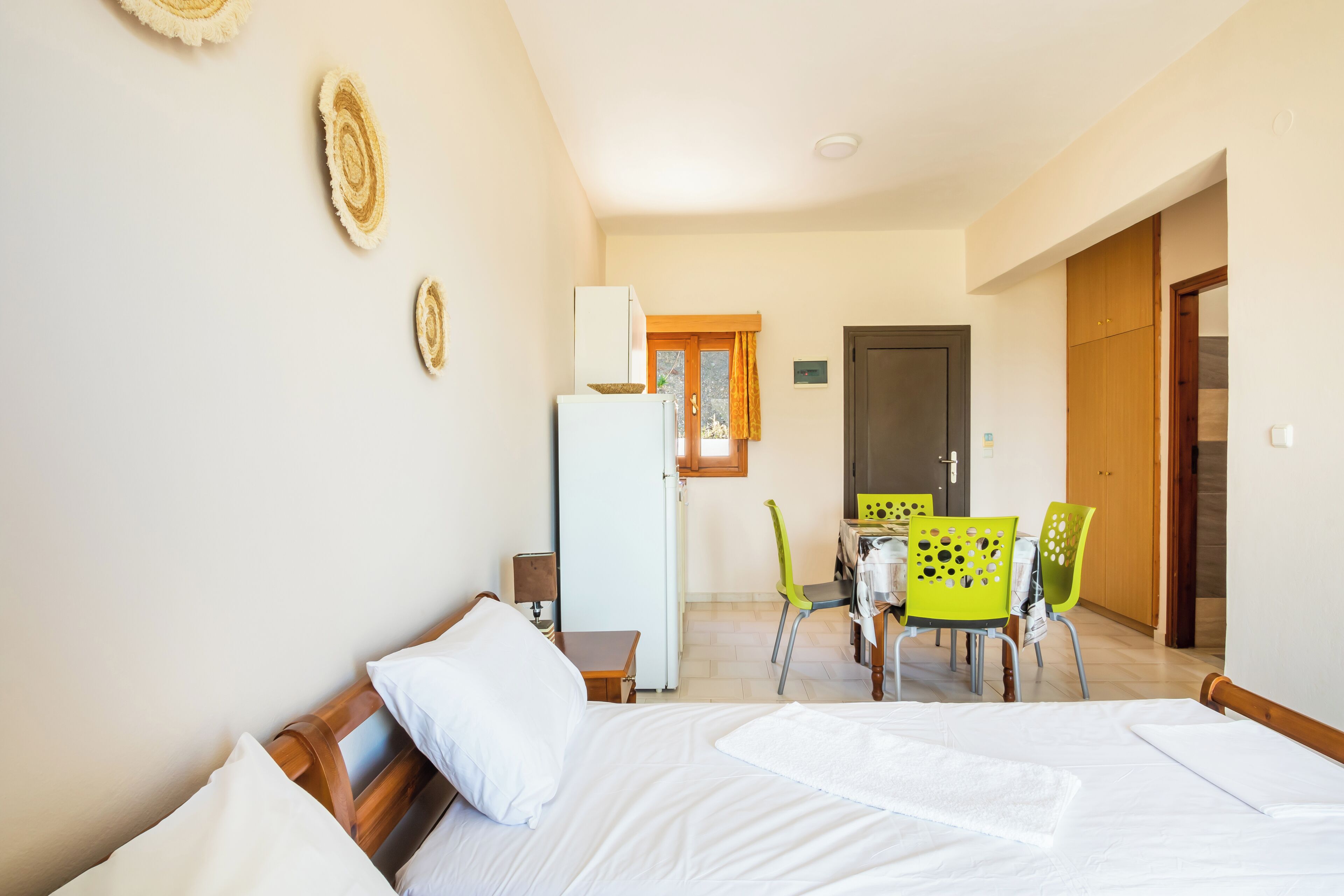 Quarto com cama, escrivaninha e cadeira. Há uma geladeira e uma porta ao fundo.