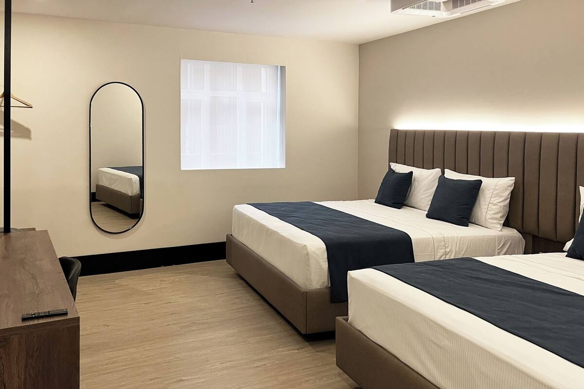 Quarto de hotel com duas camas, uma secretária de madeira, um espelho e uma janela com persianas.
