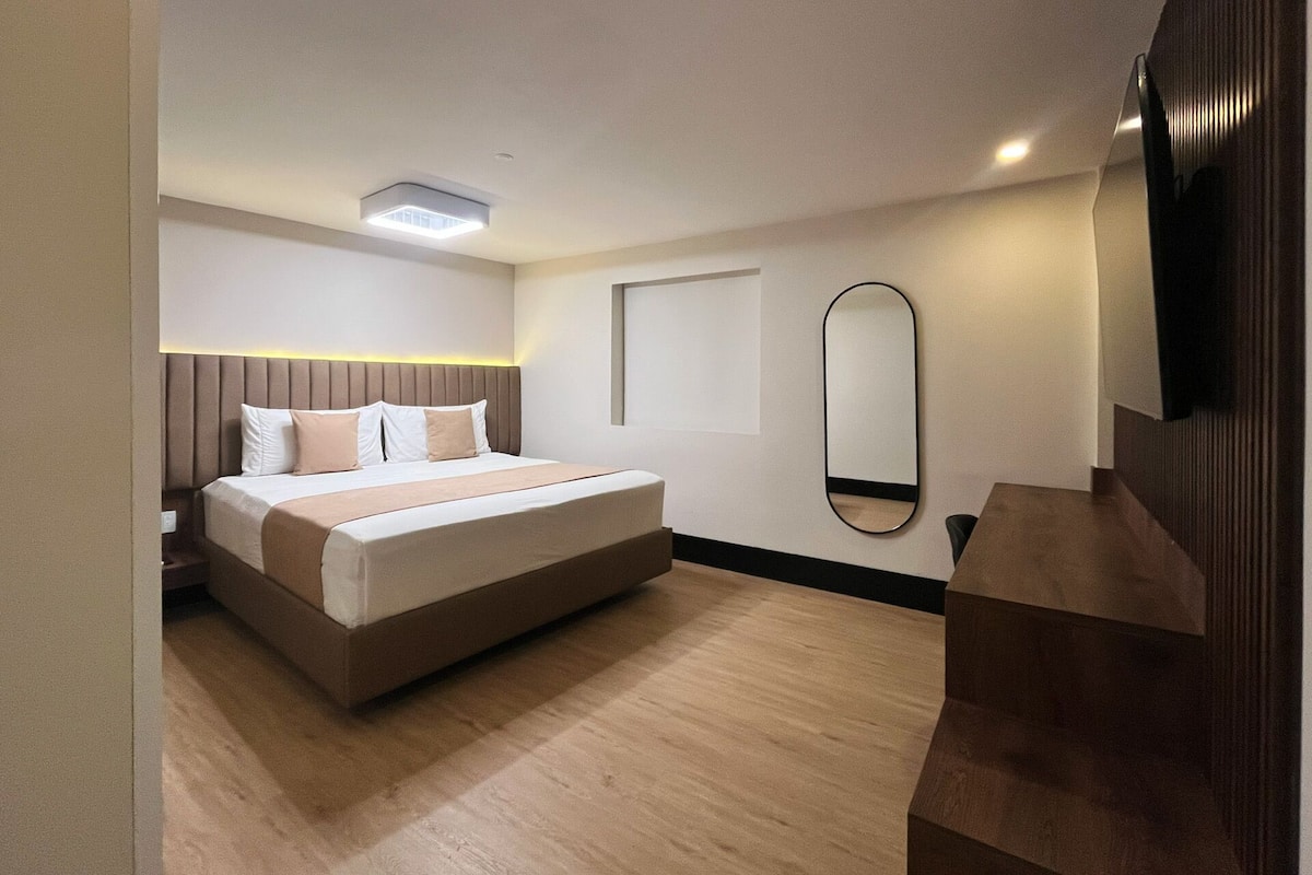 Quarto de hotel moderno com uma cama grande, uma mesinha de cabeceira, uma televisão de tela plana e uma luminária de teto.